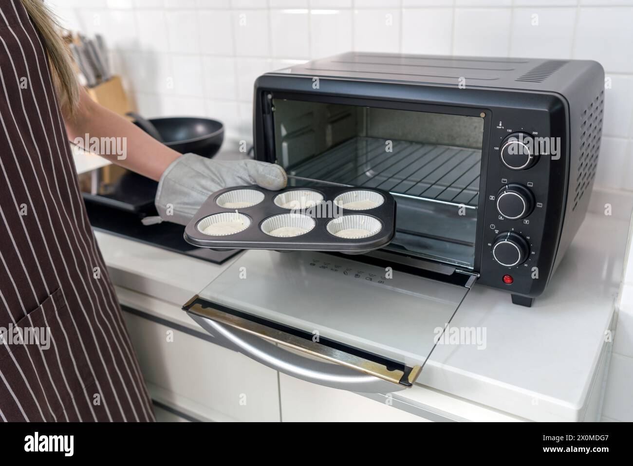 A person in an apron and oven gloves, placing a muffin tray with batter ...