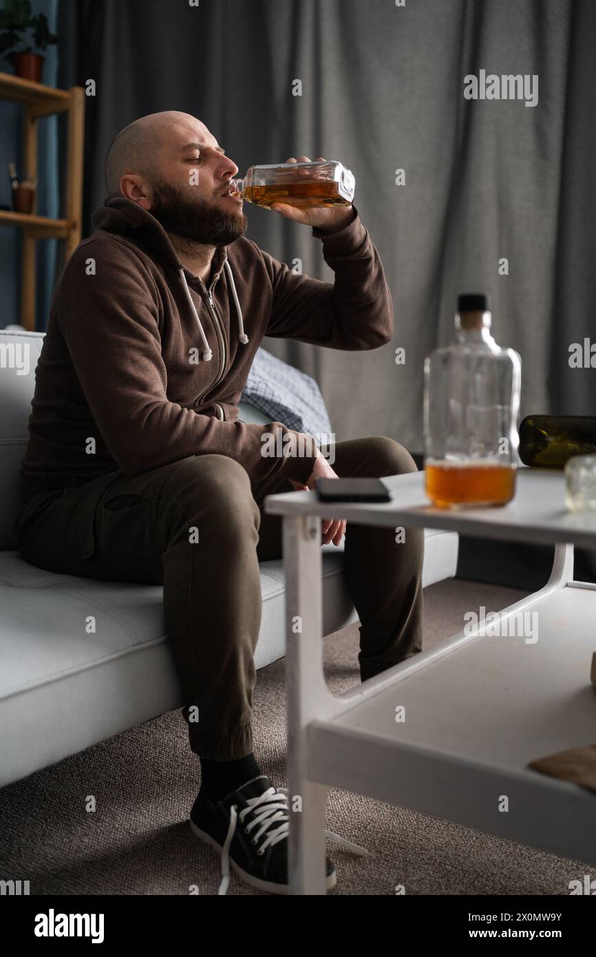 Drunk middle adult man at home on couch drinking whiskey from bottle in ...