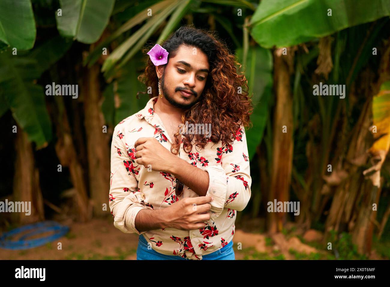 Confident gay man with curly hair, flower in hair stands in tropical ...