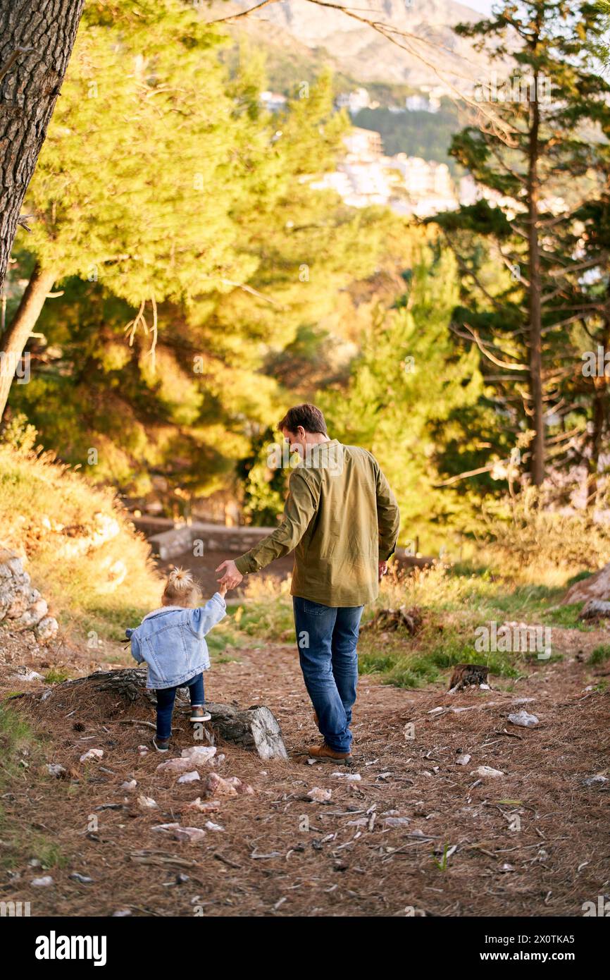 Little girl climbs a snag holding her dad hand in the forest. Back view ...