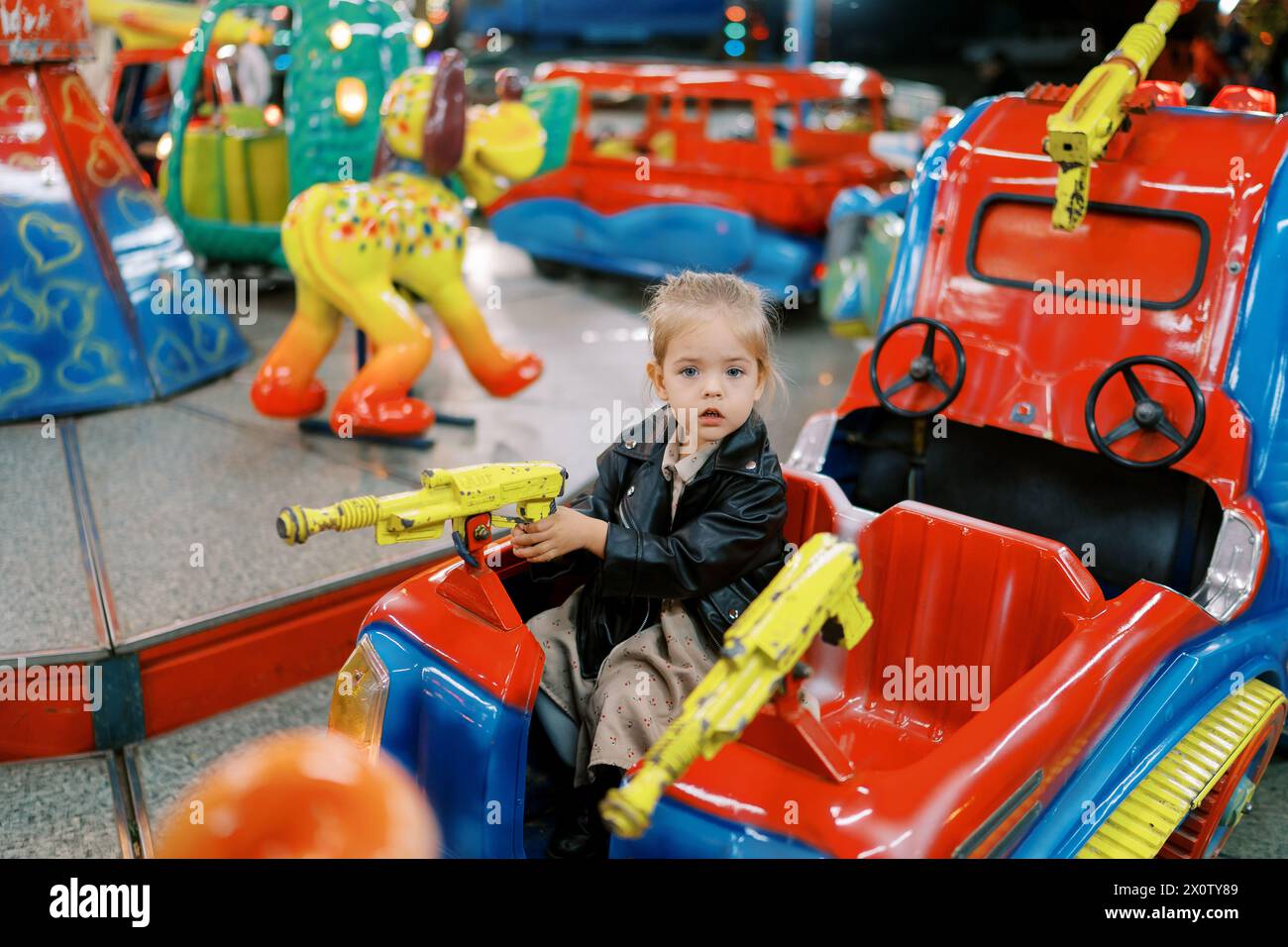 Little girl sits in a toy car on a carousel, holding the steering wheel ...