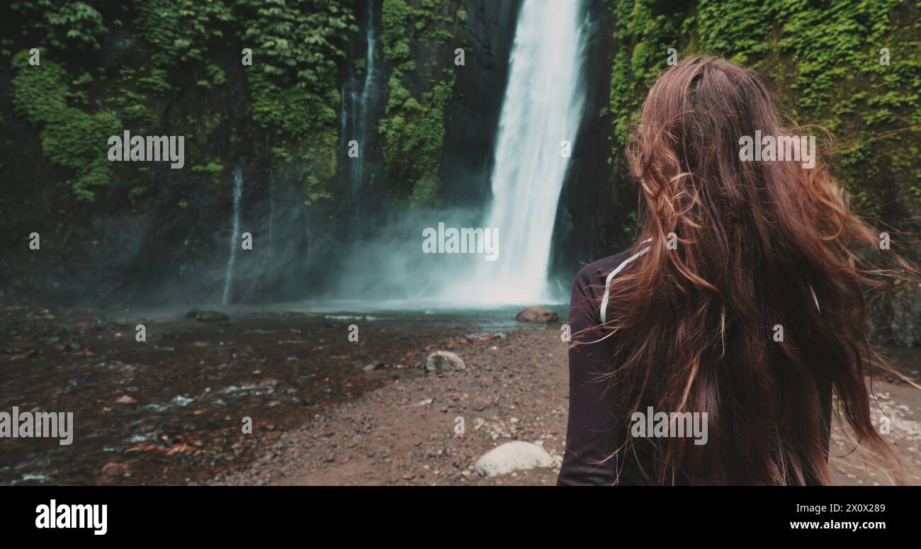 A woman stands at the base of a majestic Bali waterfall, surrounded by ...