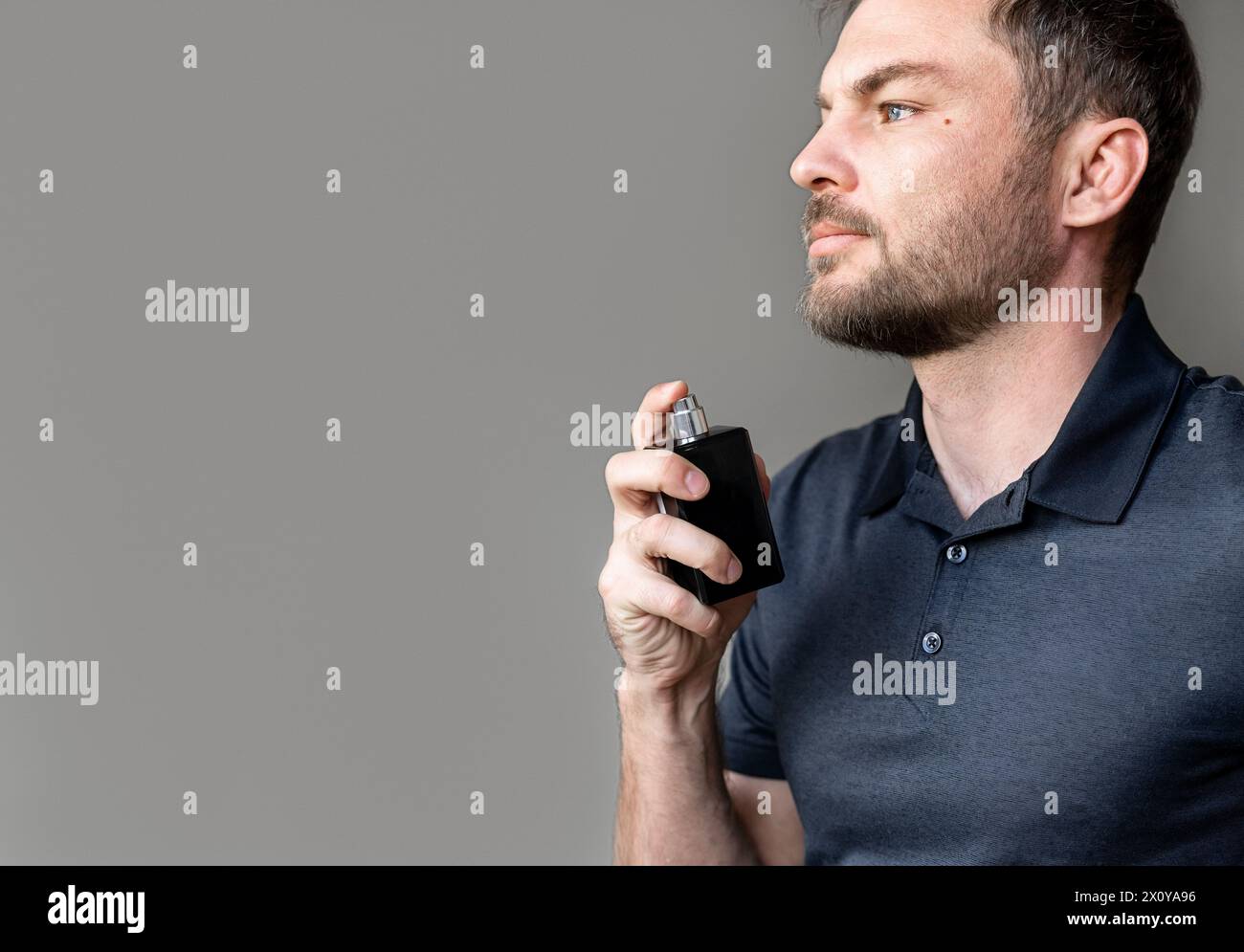 Applying perfume. Man spraying cologne on his body in front of gray ...