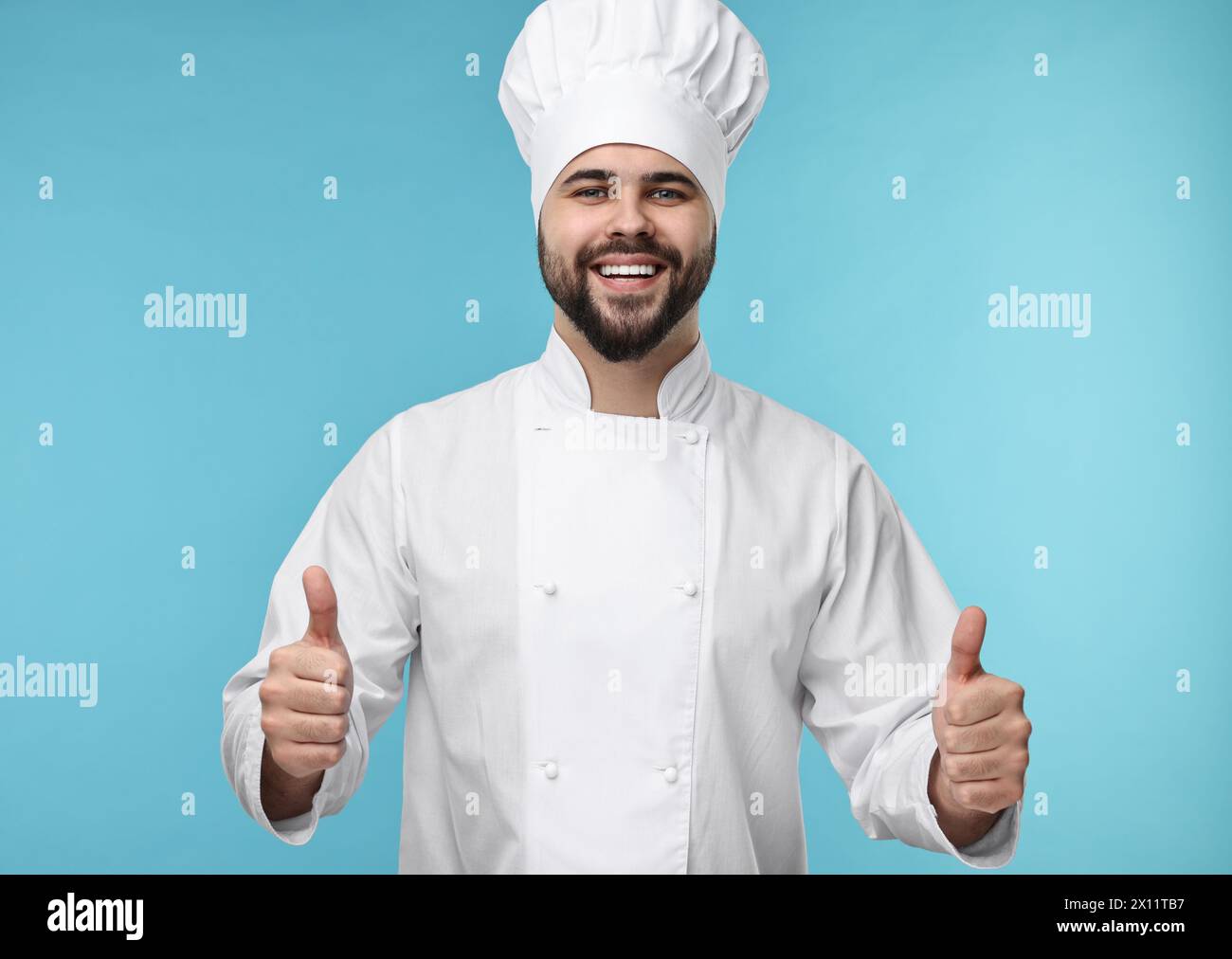 Happy young chef in uniform showing thumbs up on light blue background ...