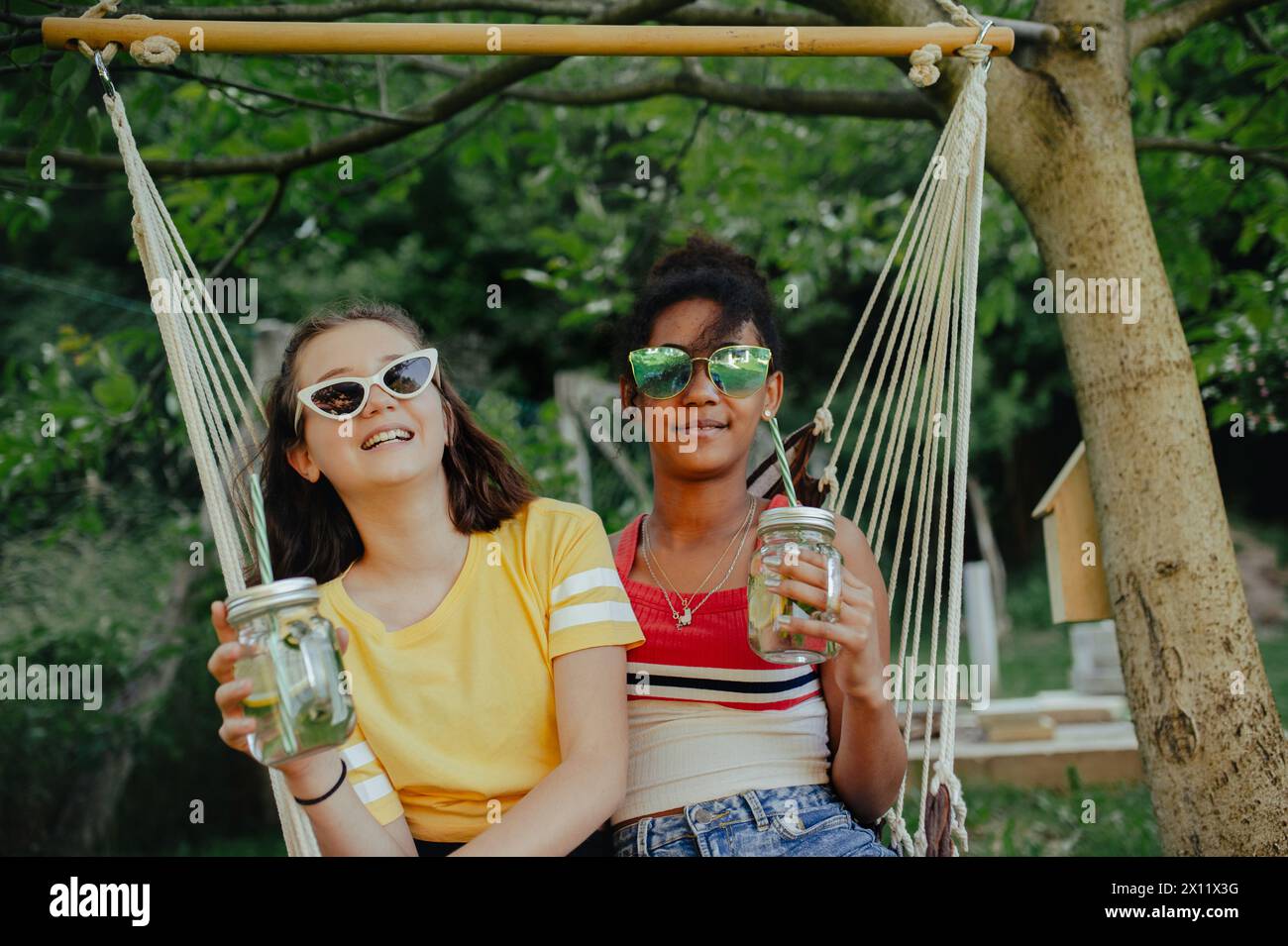 Teenage girls friends outdoors in garden, swinging on swing, drinking lemonade from jar glass ...