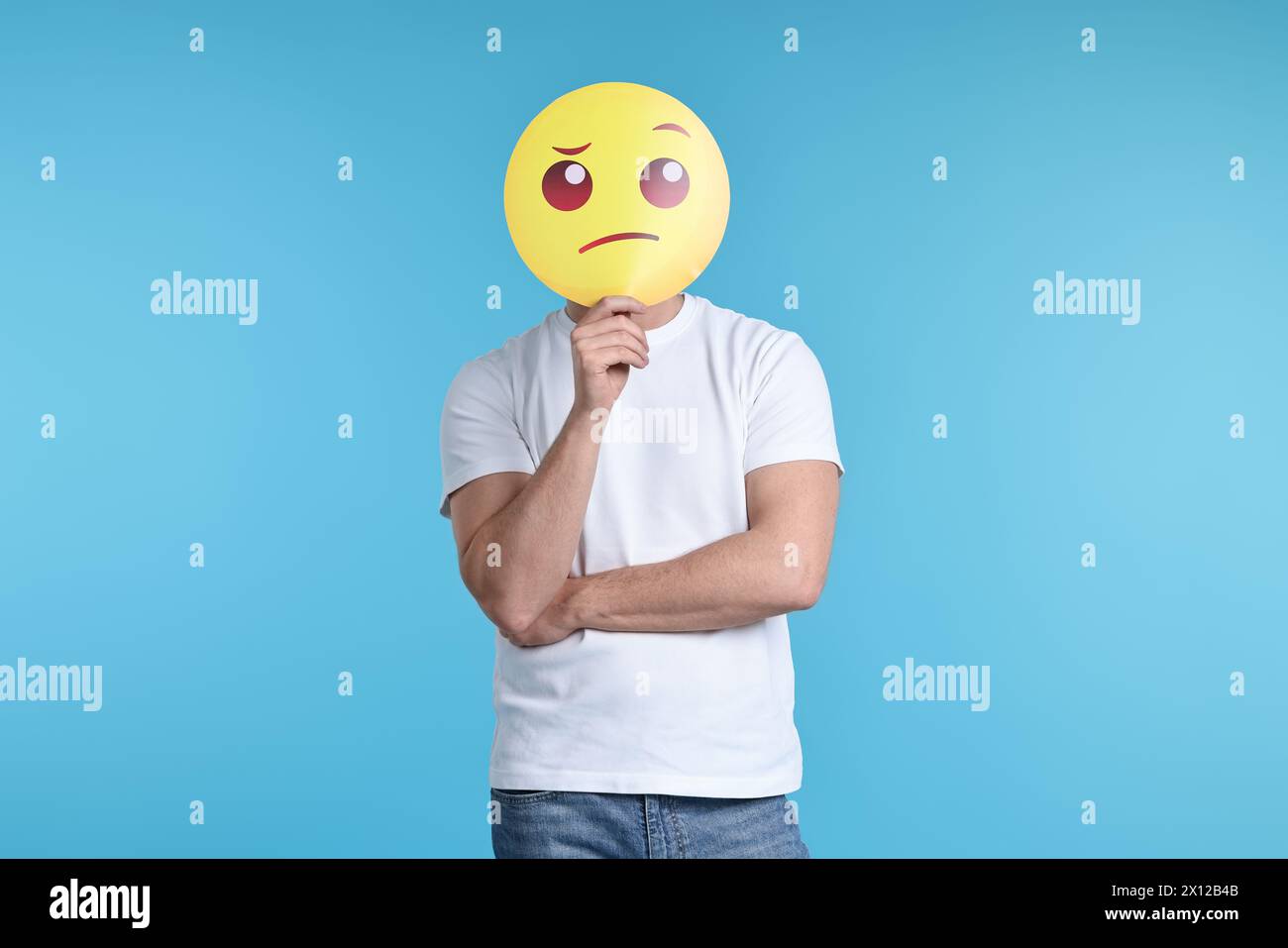 Man covering face with thinking emoticon on light blue background Stock Photo - Alamy