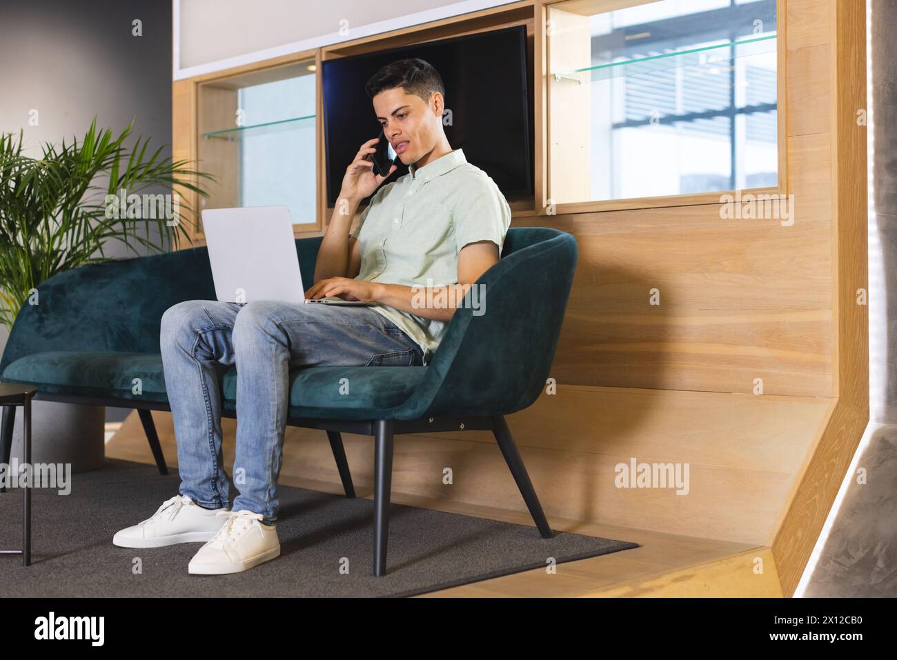 A biracial man talking on phone, holding laptop on his lap in a modern ...