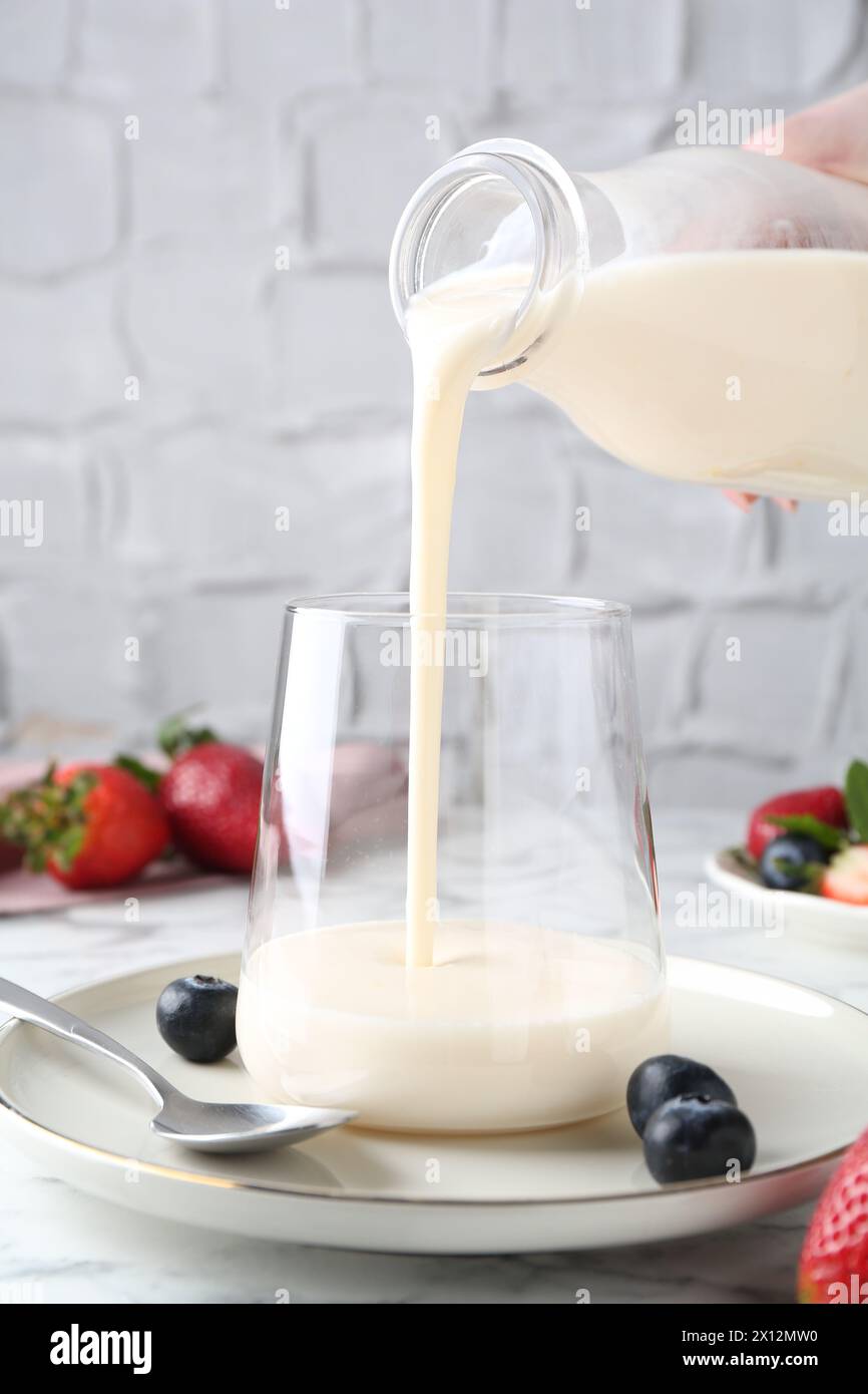 Woman pouring tasty yogurt into glass at white marble table, closeup ...