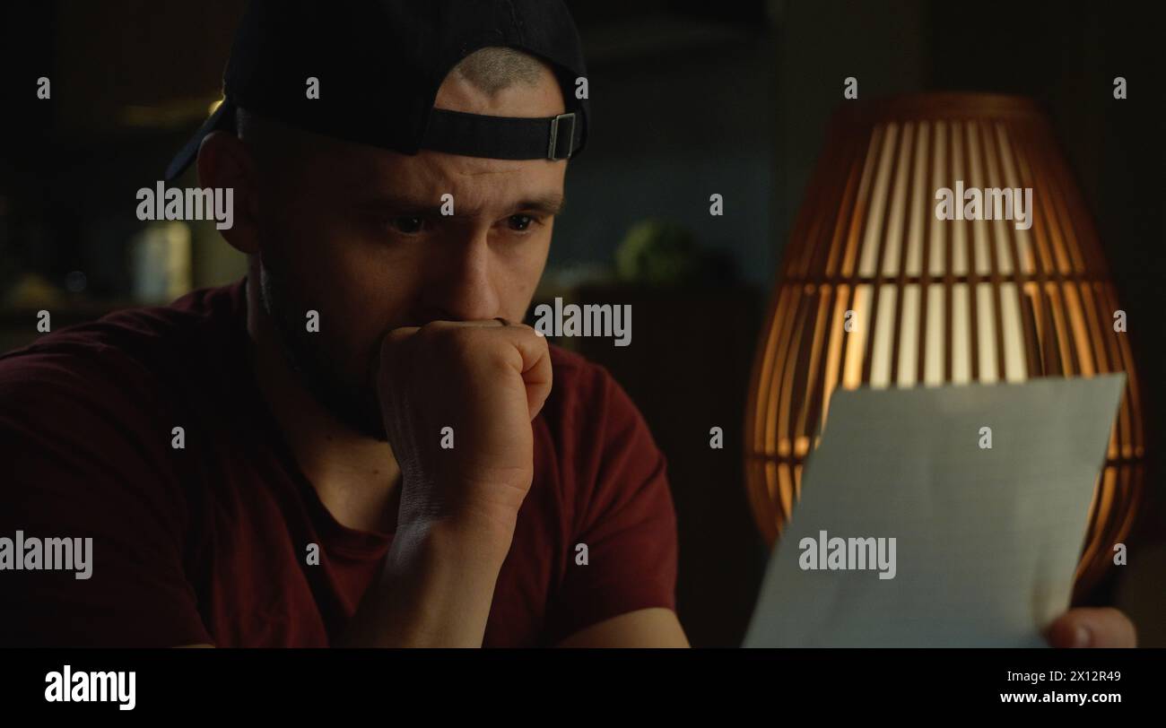 Shocked man reading paper document sitting iin dark room at home ...