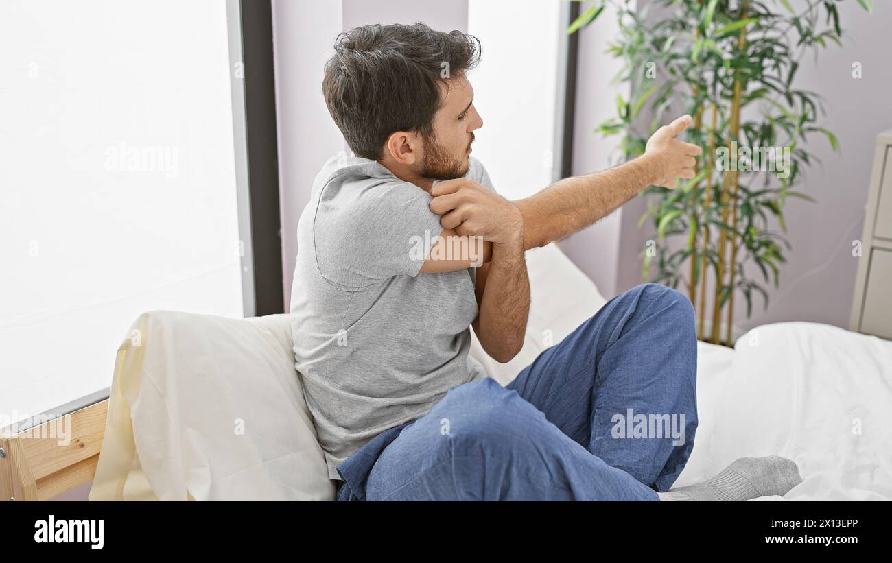 A Young Hispanic Man With A Beard Sits On A Bed Stretching His Arm In A