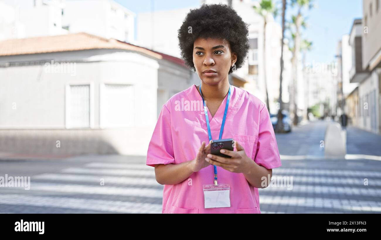 African american nurse woman with curly hair in pink scrubs using smartphone on city street ...