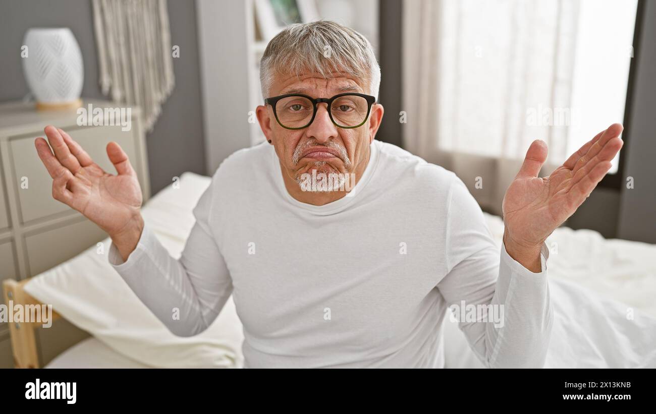 Confused senior man with grey hair and glasses sitting on a bed in a ...