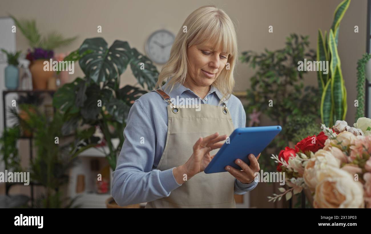 A blonde woman florist in an apron uses a tablet inside a vibrant ...