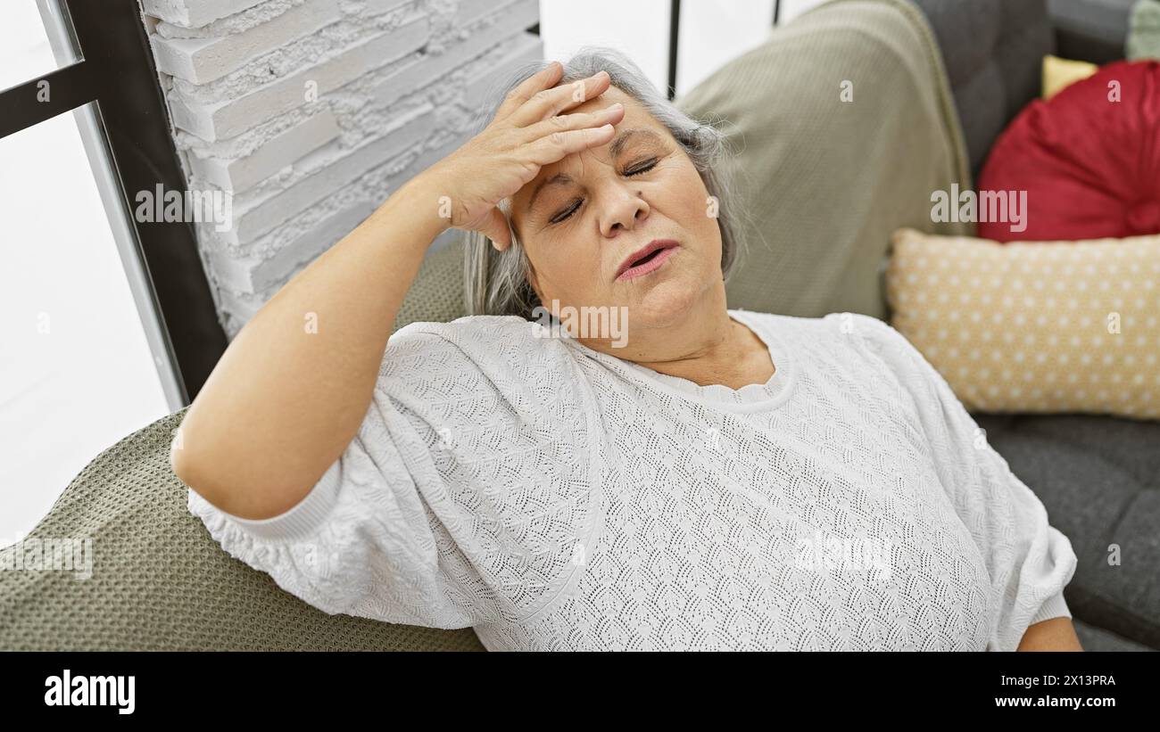 A senior woman with grey hair looks distressed while sitting on a couch ...