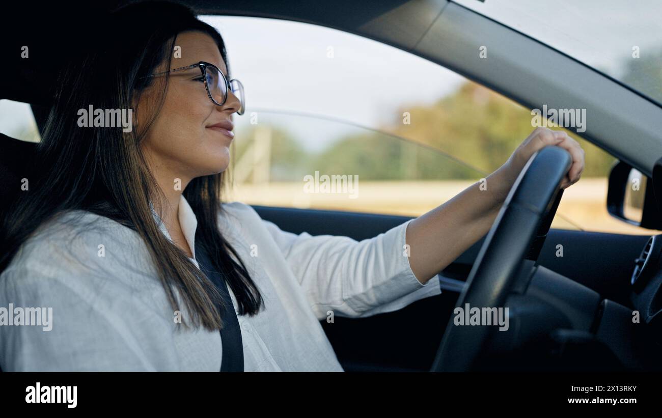 Young beautiful hispanic woman driving a car smiling wearing glasses on ...
