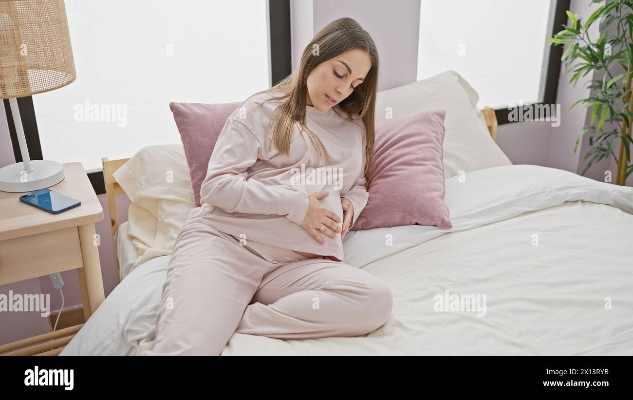 A young hispanic pregnant woman in pajamas gently touching her belly ...