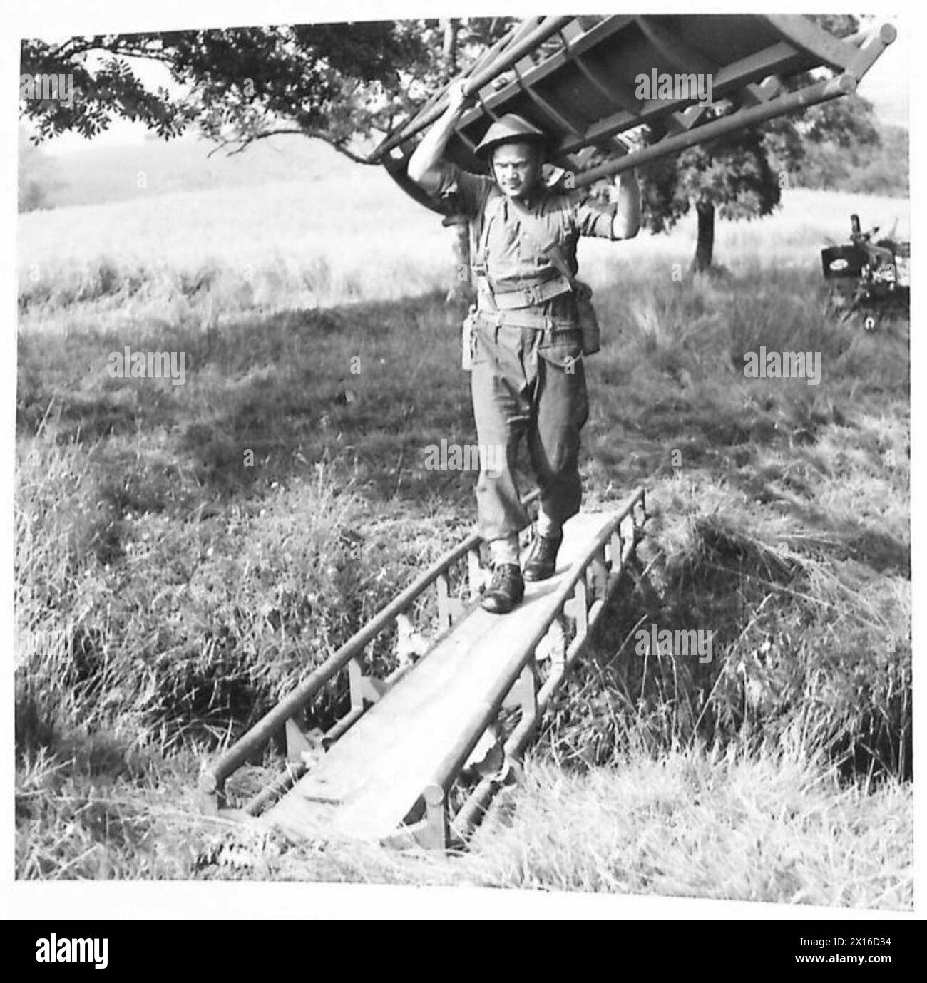 A man carries one section of a bridge across a stream while another ...