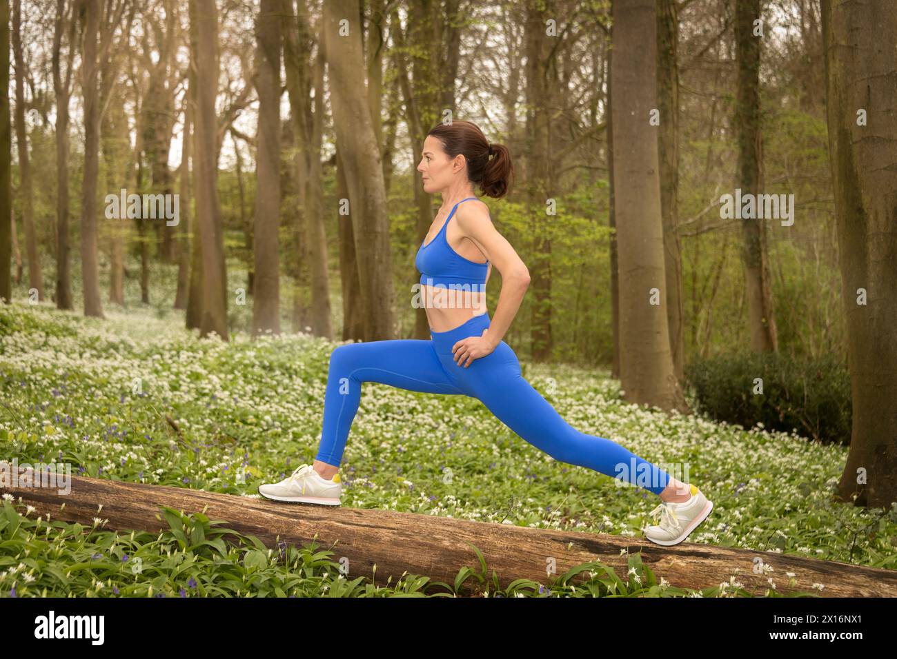 Sporty woman doing stretching exercises on a log in a forest, wild garlic and flowers background ...