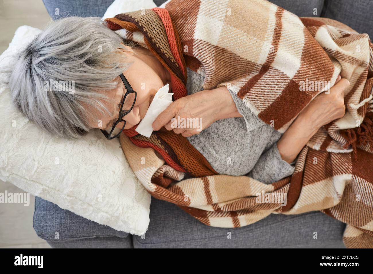 Senior sick woman lying on sofa at home wrapped in a blanket suffering ...