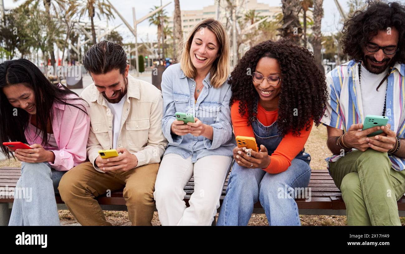 Multiethnic group of people sitting in a row outside, using phones ...
