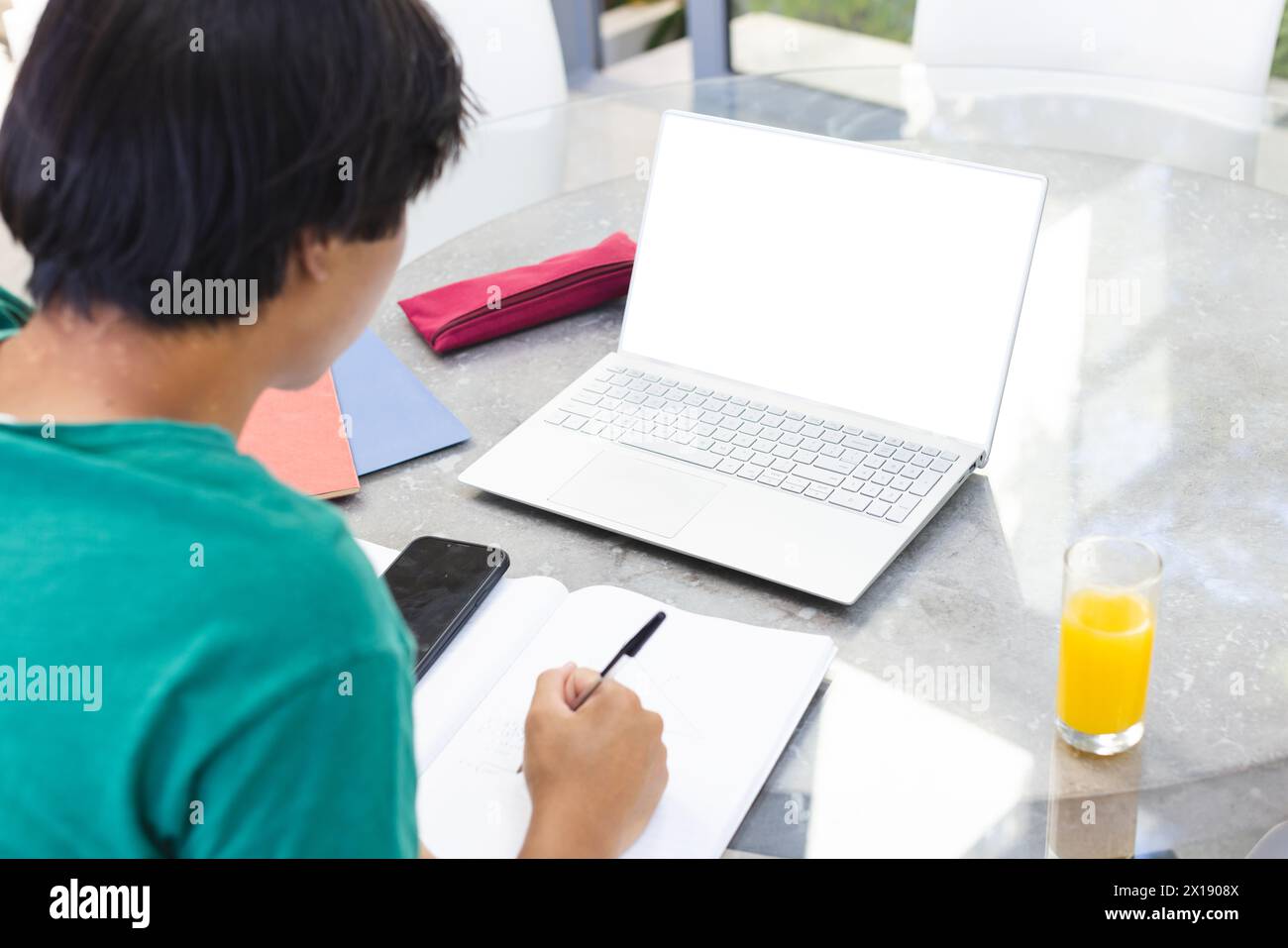 At home, outside, teenage Asian boy writing in notebook, laptop open ...
