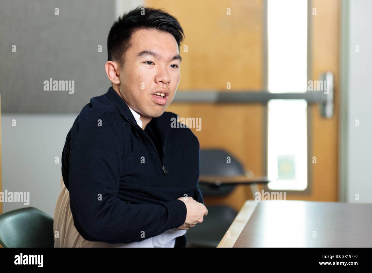 A young Asian male student in a classroom scene putting on a sweater ...