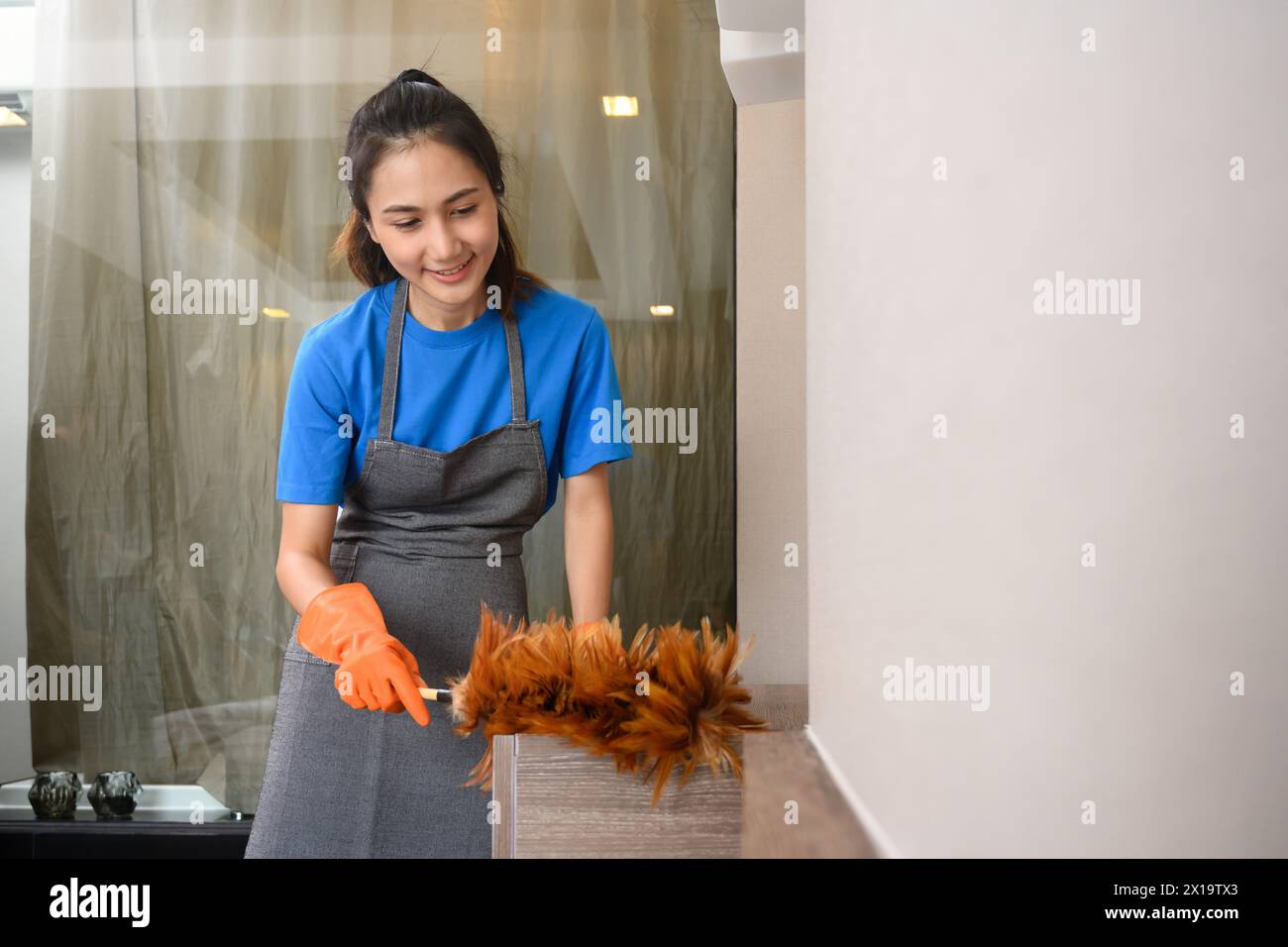 Smiling female cleaner wearing apron dusting in bedroom. Cleaning ...
