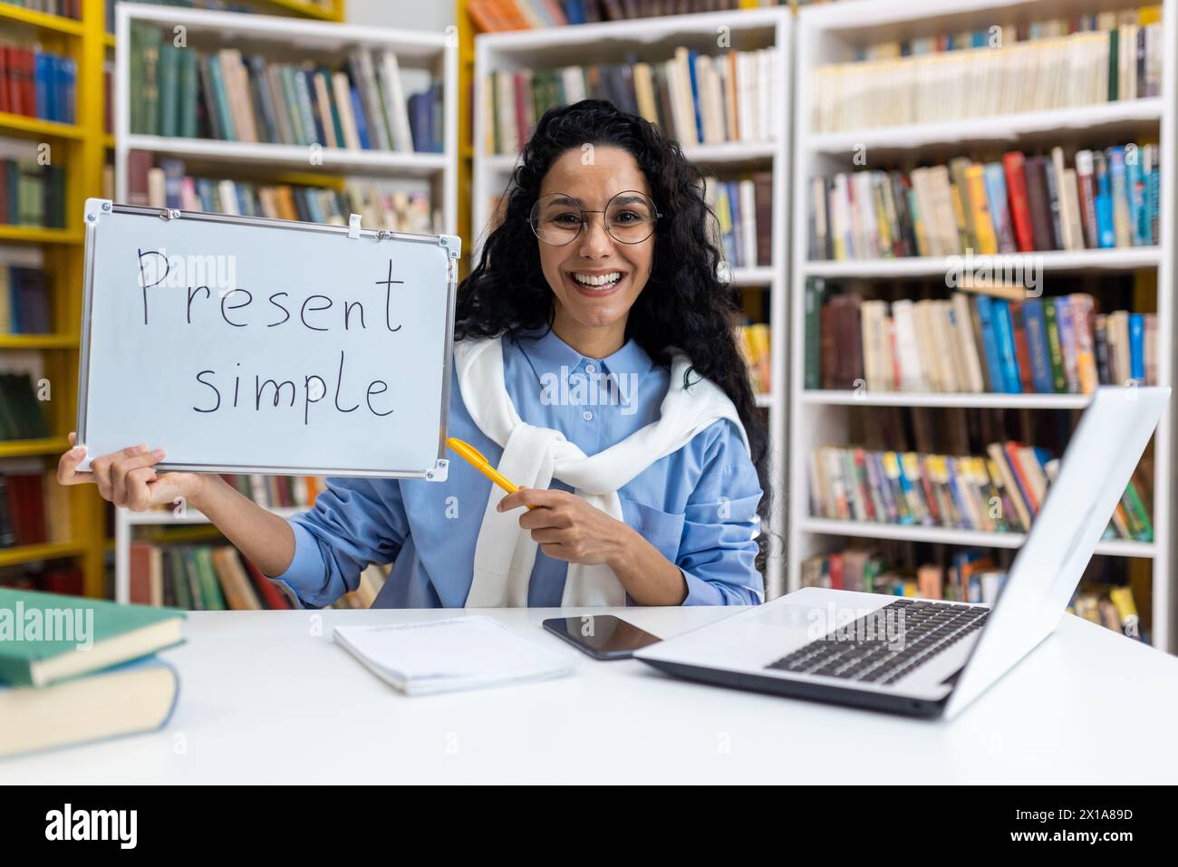 Cheerful female teacher holding a board with 'Present Simple' written on it, demonstrating ...