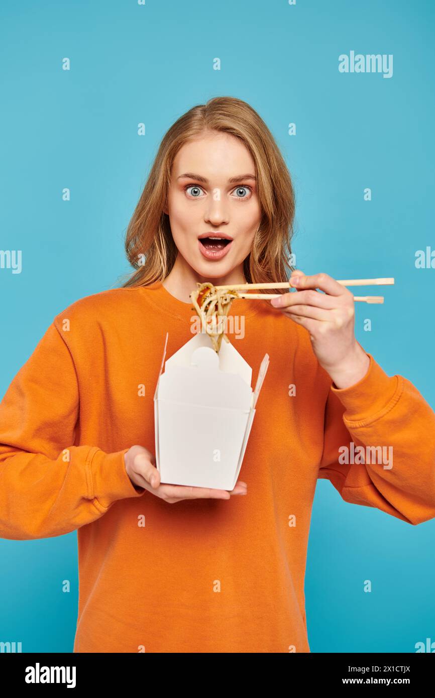 A chic blonde woman elegantly holds chopsticks and a box of noodles ...