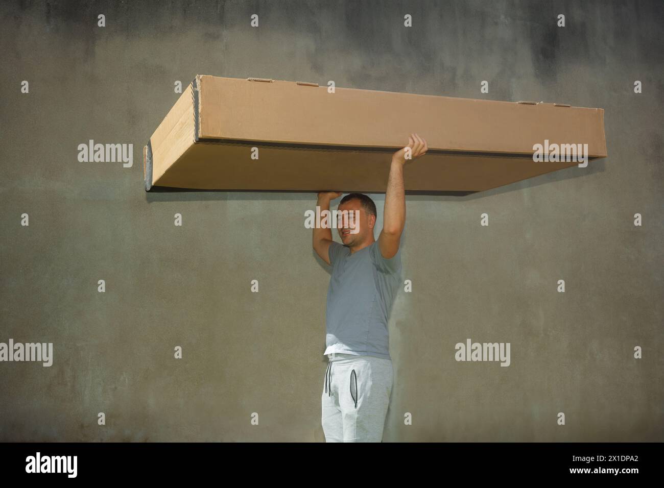 Man lifts a large cardboard box over his head against grey wall Stock ...