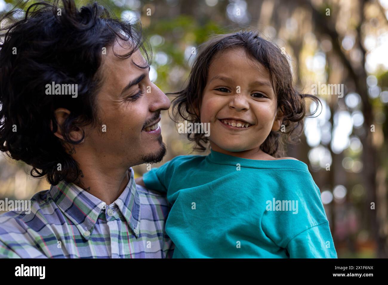 Latin American dad carries his 3 year old son in his arms in the park ...