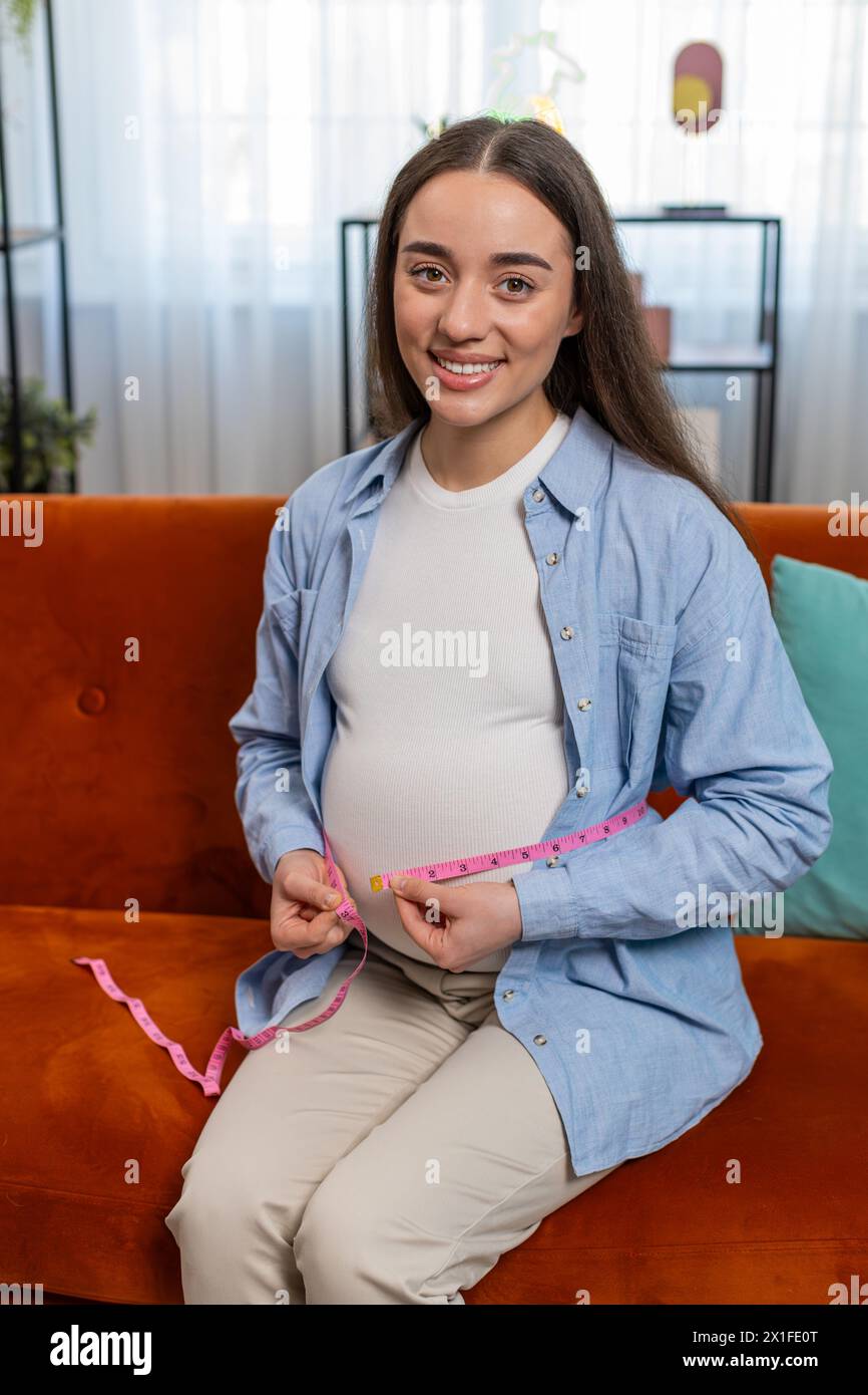 Smiling pregnant woman on couch measuring belly with measure tape ...
