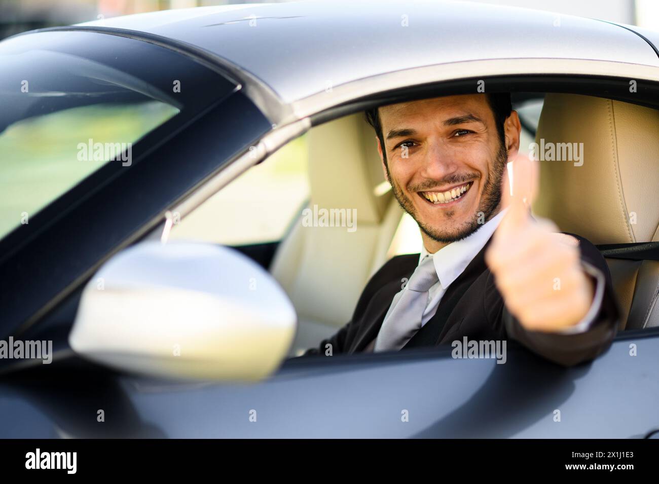 Smiling young businessman in a suit gives a thumbs up from the driver's ...