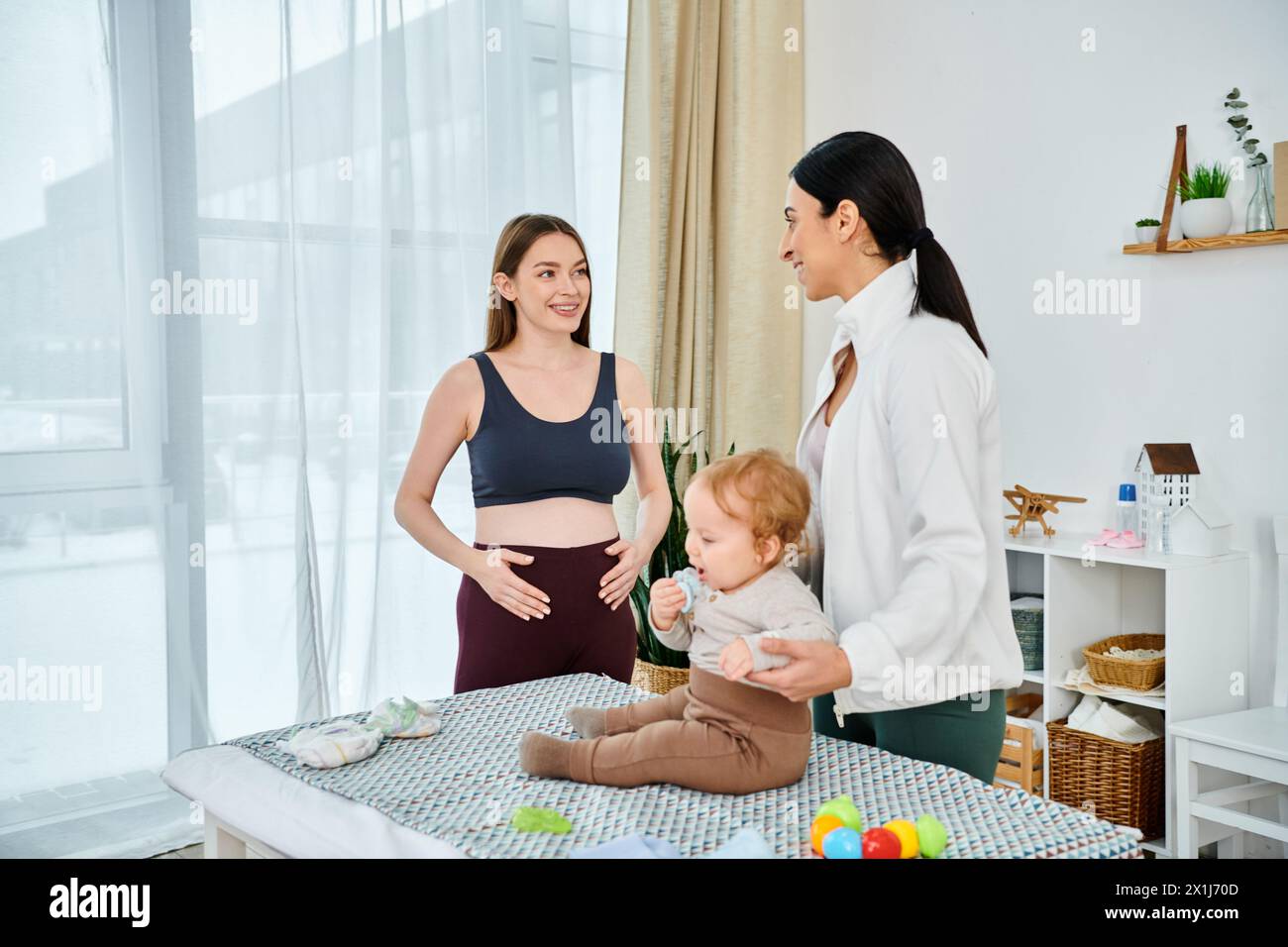 A young beautiful mother stands next to her baby in a cozy room ...