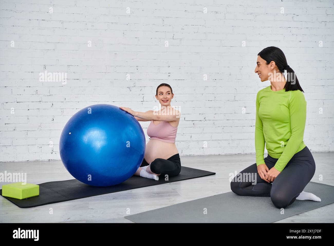 Two women, one pregnant, are seated on yoga mats engaging with a large exercise ball under the ...