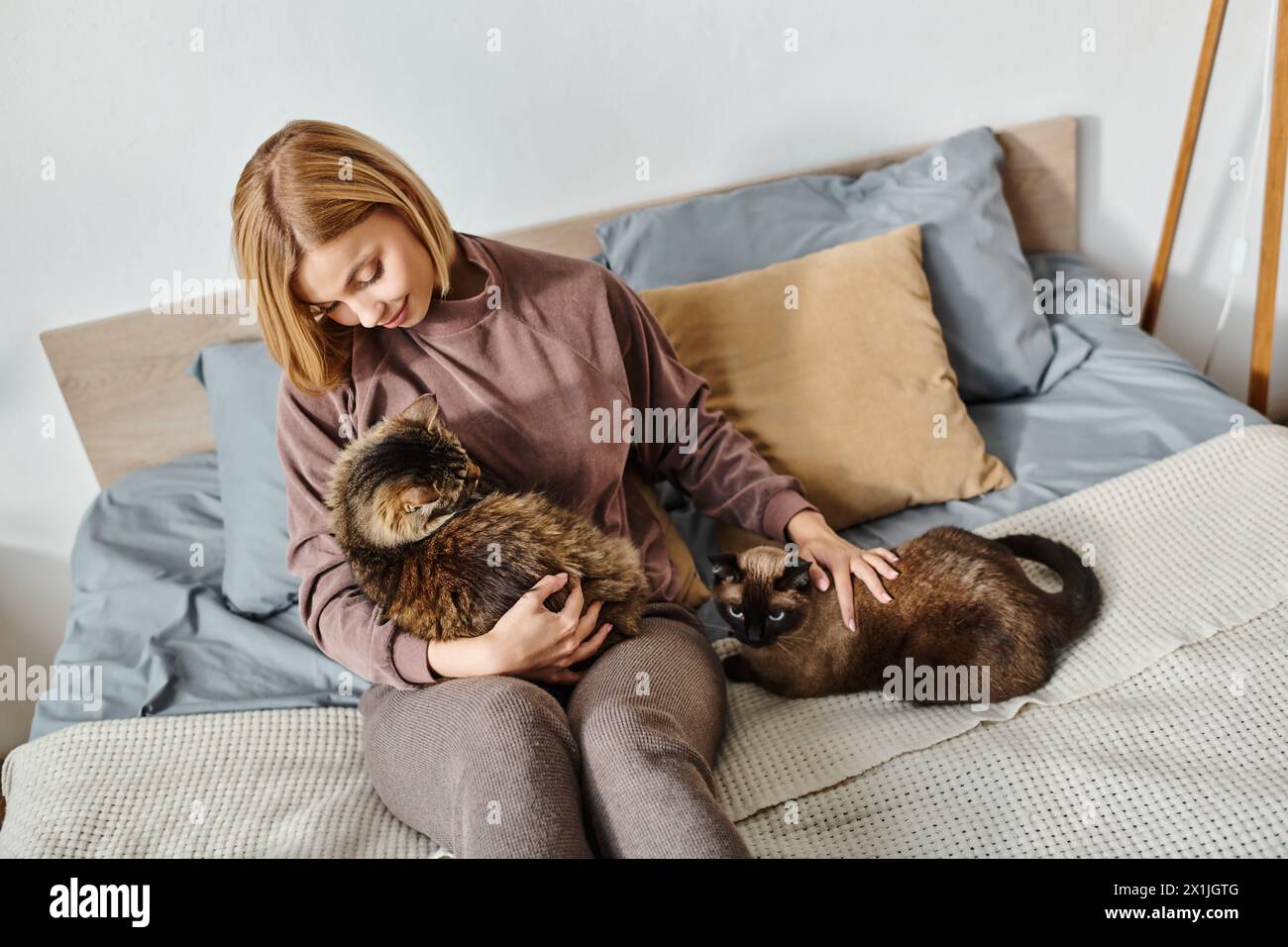 A woman with short hair peacefully sitting on a bed, holding two cats ...