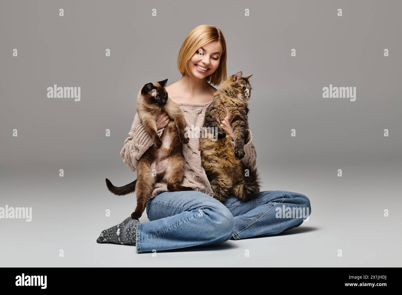 A woman with short hair sitting on the floor, holding two cats closely ...