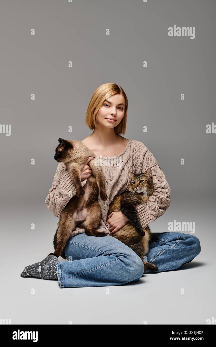 A short-haired woman sits on the floor, cradling two cats in her arms ...
