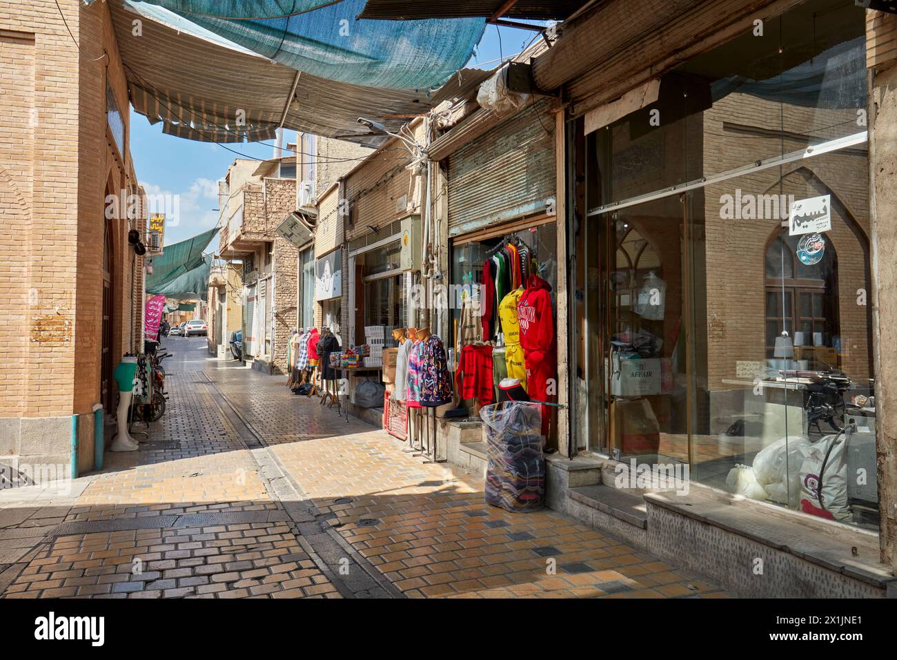 A narrow cobbled street with sun shade canopy lined with small shops in ...