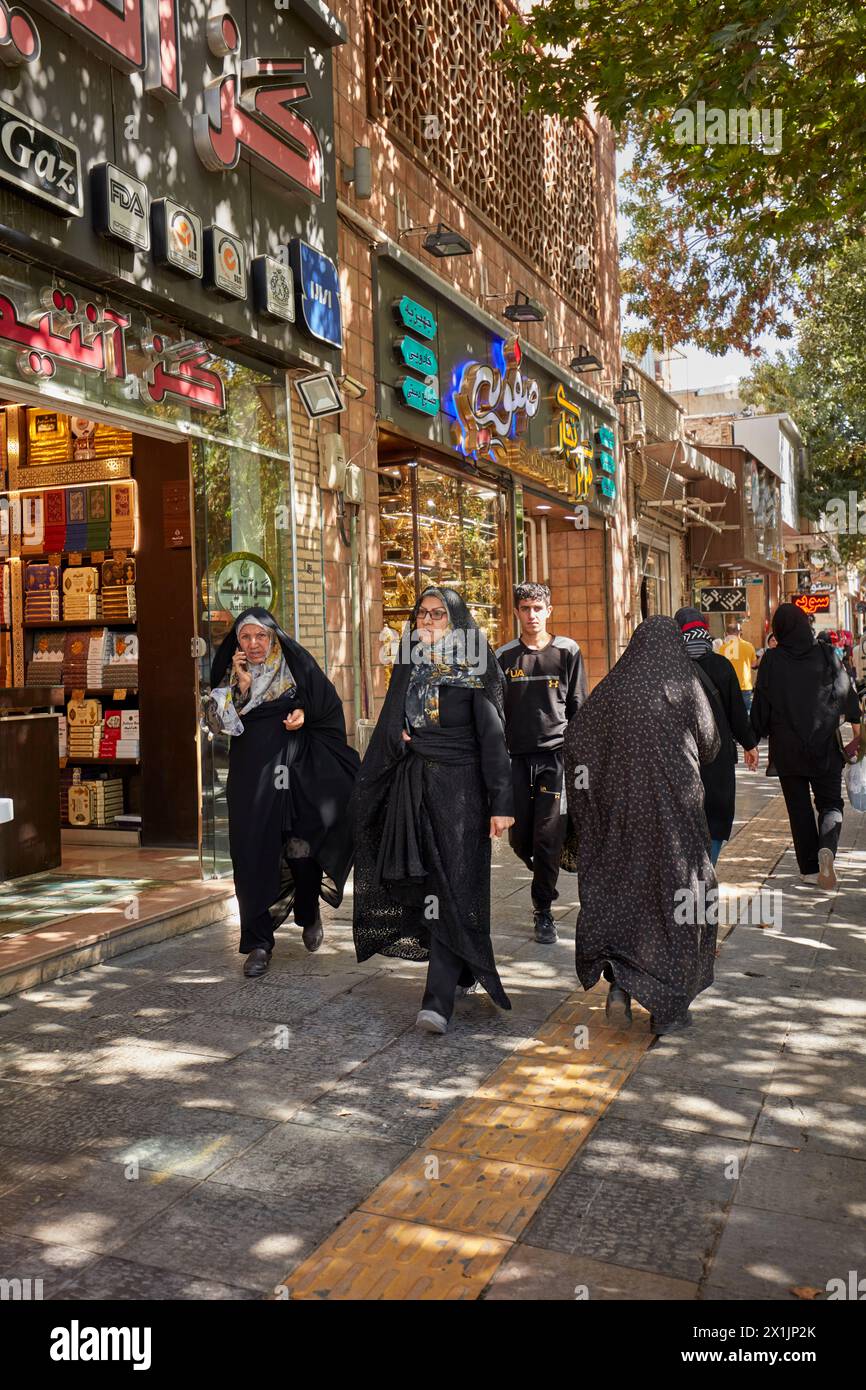 Iranian women wearing traditional black chadors walk along a narrow ...