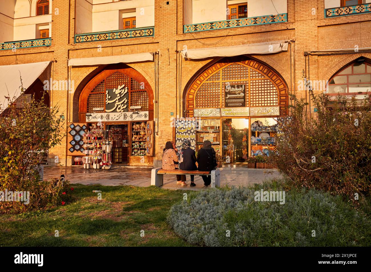 Three Iranian women sit on a bench in front of a handicraft shop window ...