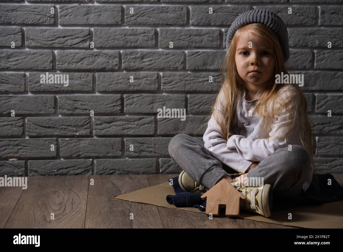 Homeless little girl with figure of house sitting near grey brick wall ...