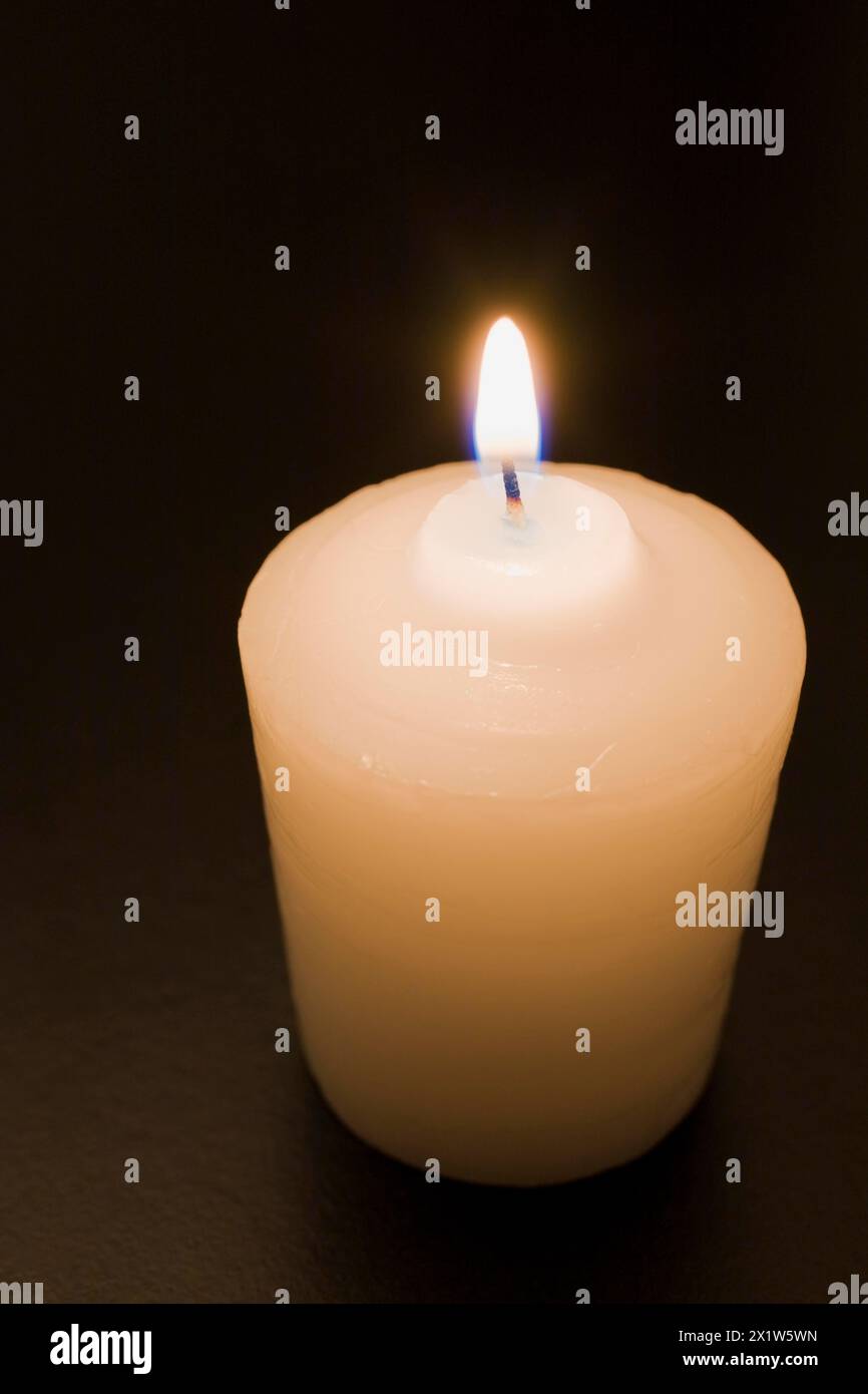 Close-up of white wax candle with bright lit flame on black background ...