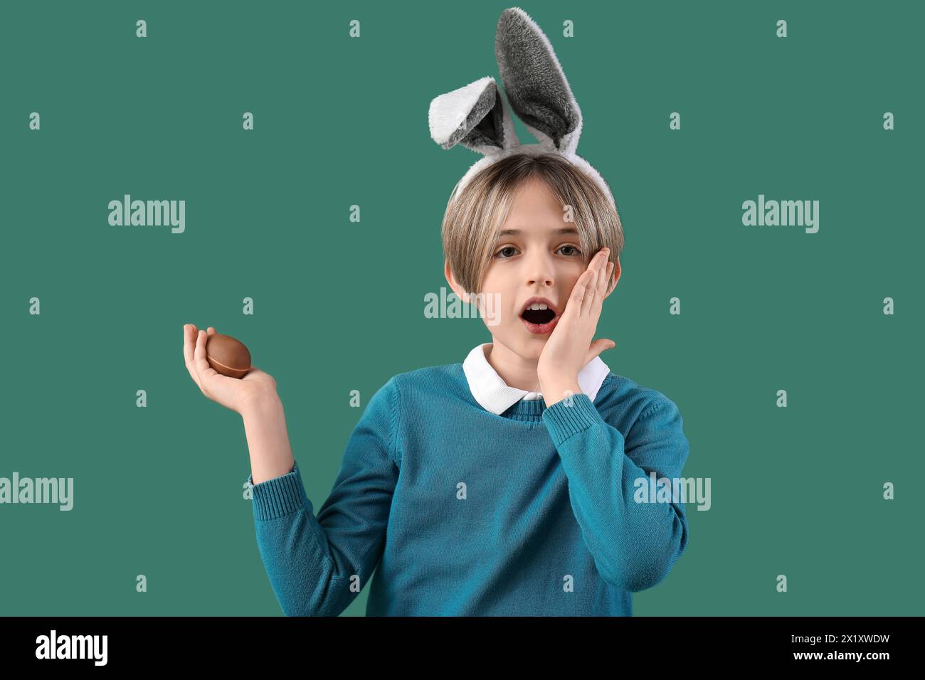 Shocked little boy in bunny ears with chocolate egg on green background ...