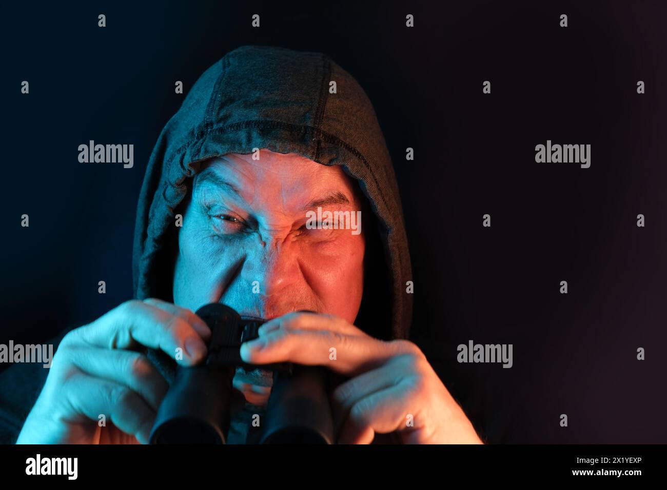 middle-aged man, stalker in hood looks through black binoculars in dark ...