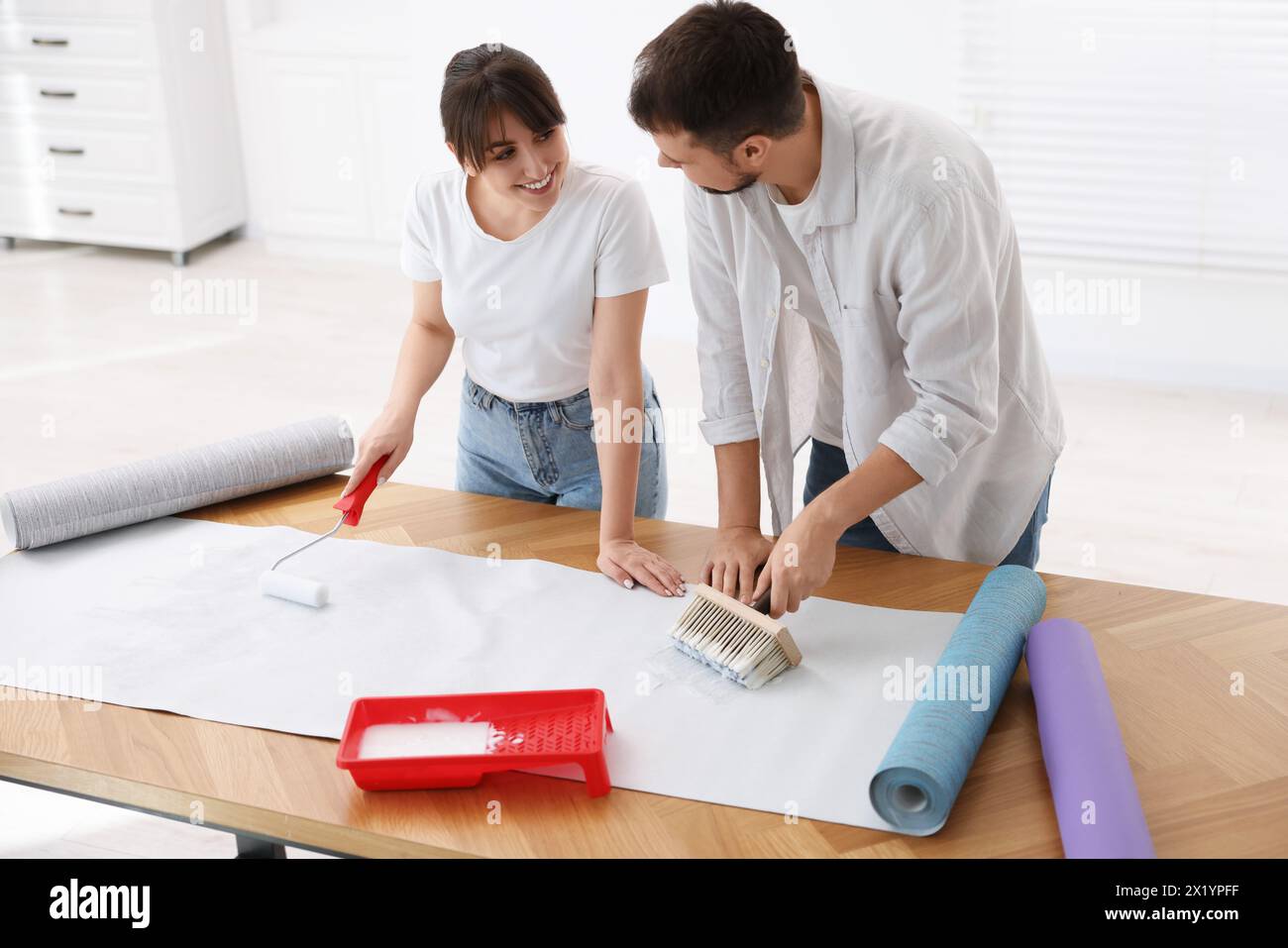 Woman and man applying glue onto wallpaper sheet at wooden table ...