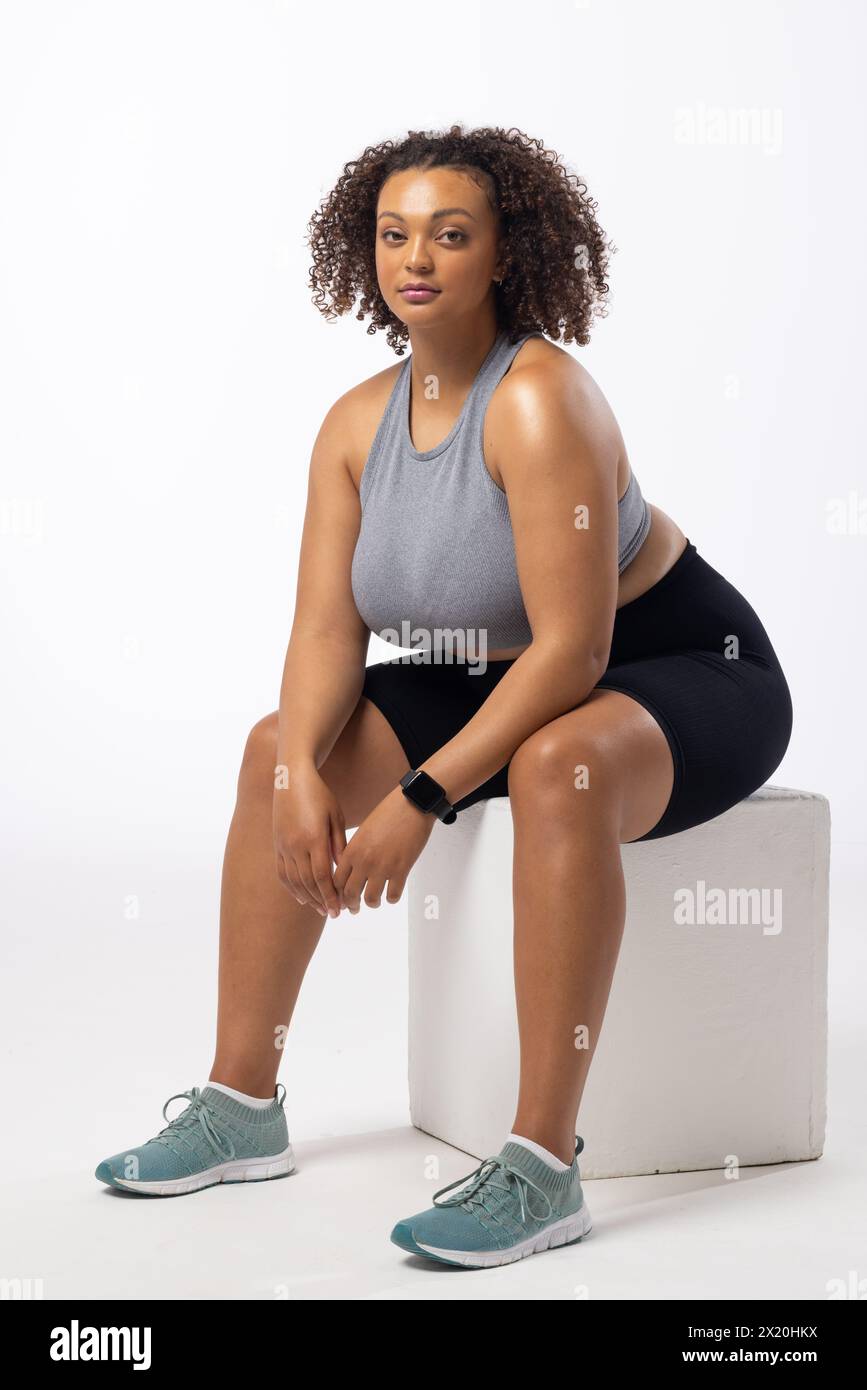 Biracial young female plus size model sits on white block, white ...