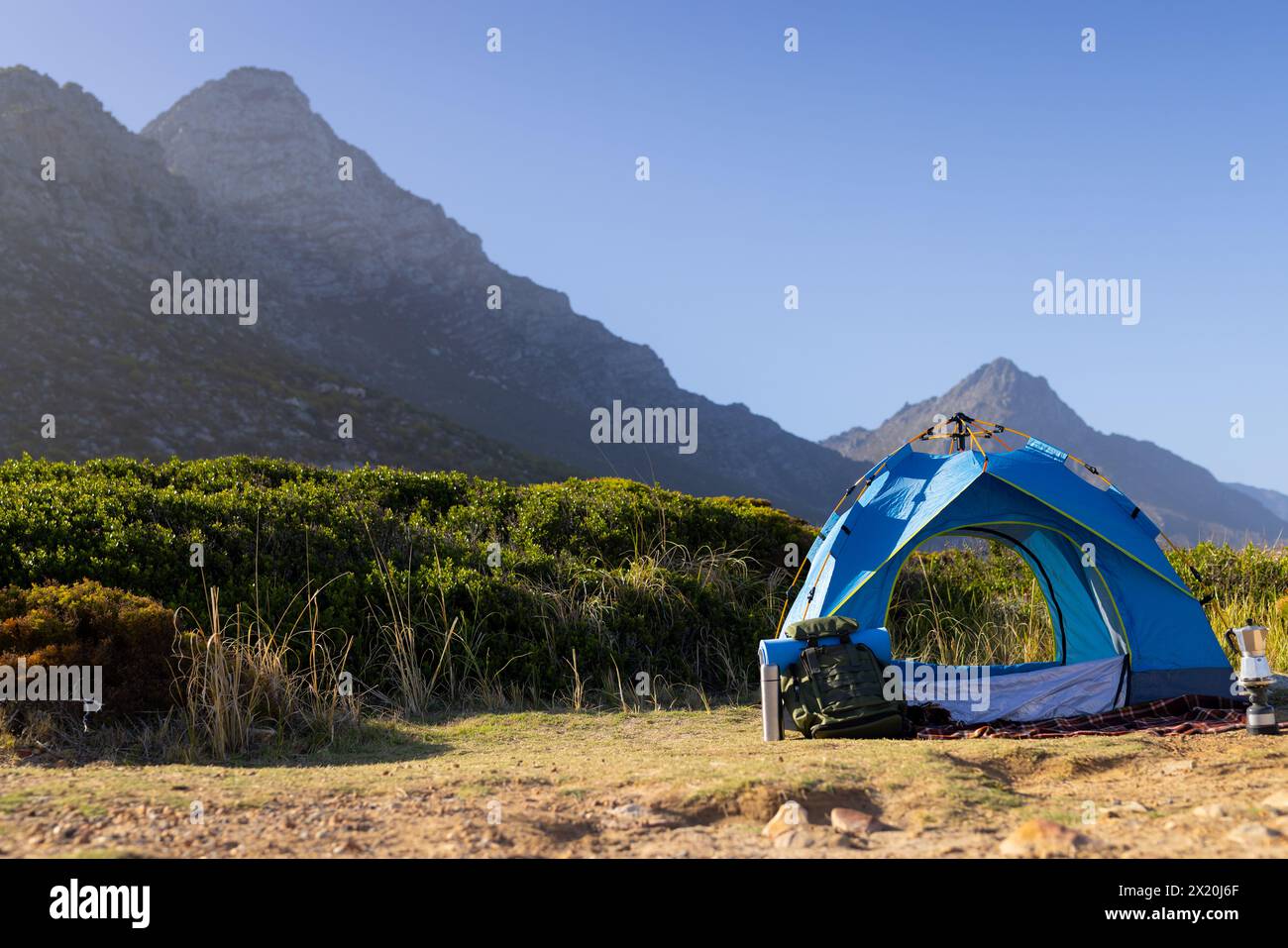 A blue tent sits at the foot of tall mountains, surrounded by greenery ...