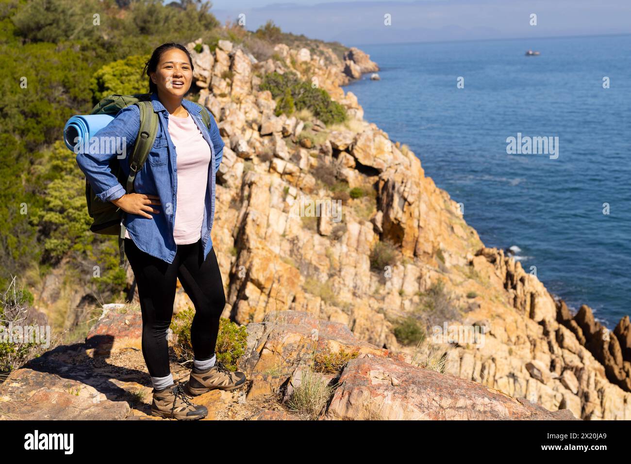 Biracial female hiker standing on rocky terrain, overlooking the sea ...