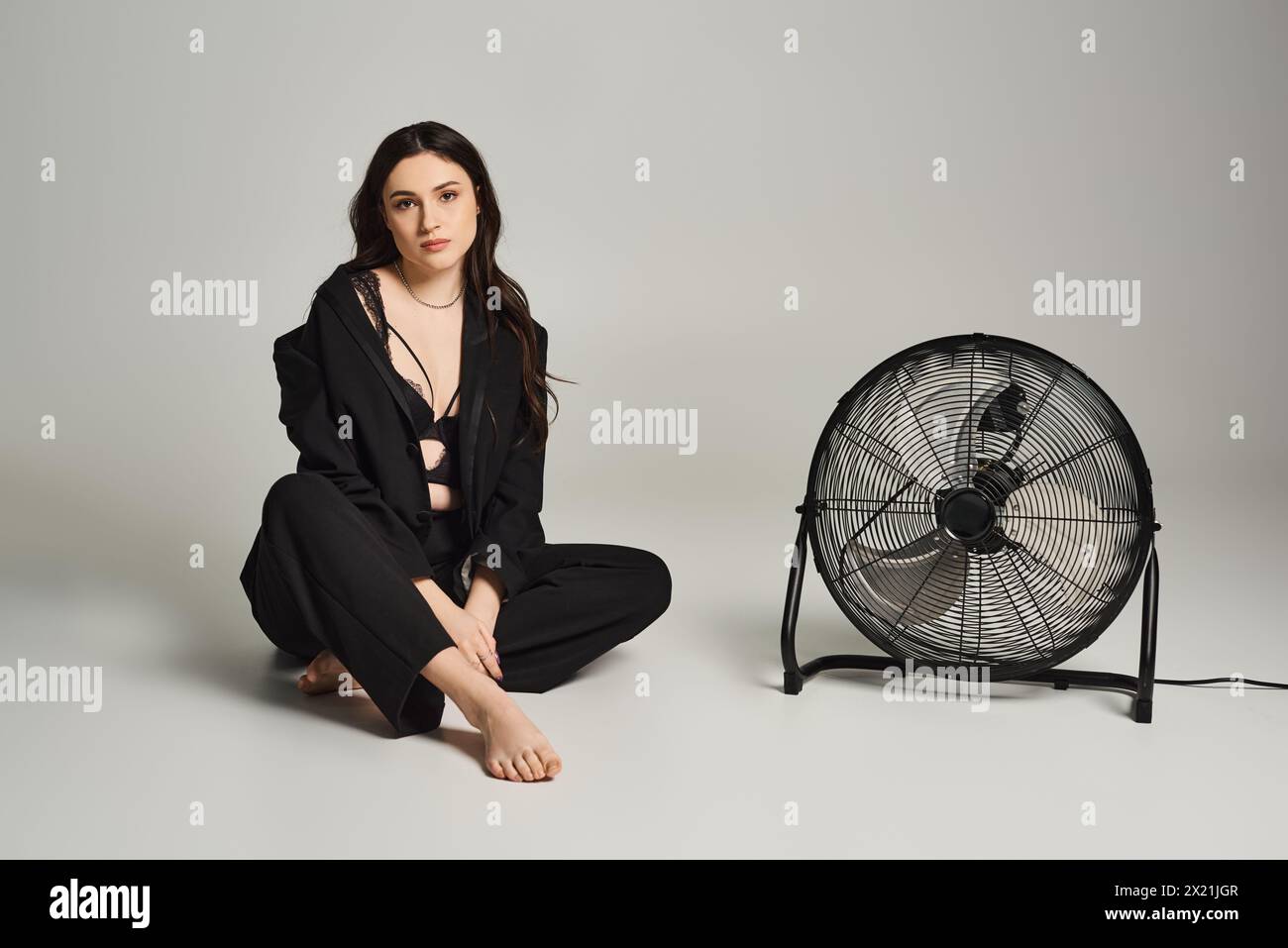 A beautiful plus-size woman in stylish attire sits gracefully next to a spinning fan on a gray ...